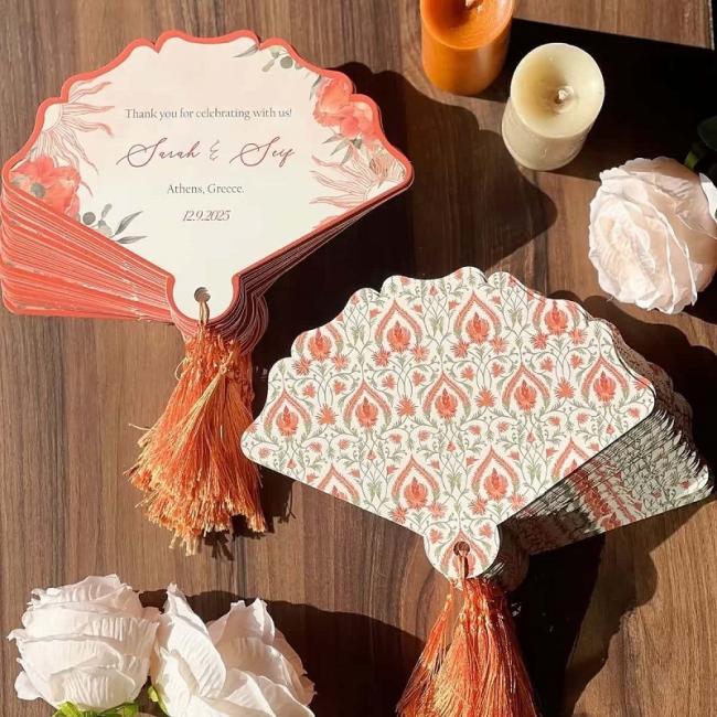 Terracotta and patterned vintage-style fan wedding invitation cards arranged with bright orange tassels on a wooden tabletop