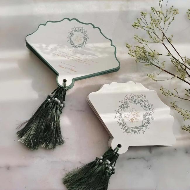 Flat lay of a green-bordered scalloped fan wedding invitation card featuring gold foil monogram crest and a deep green silk tassel