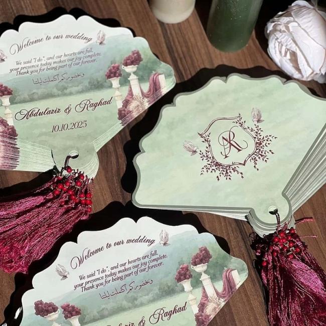 Assorted wedding invitation fan cards in sage green and burgundy featuring watercolor venue illustrations, monograms, and beaded tassels
