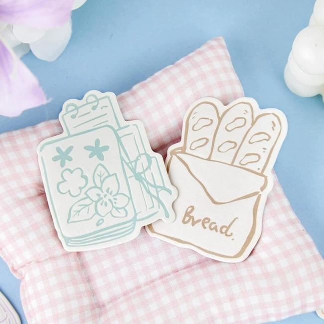 Close-up shot of memo pad and bread bag die-cut scented cards resting on a pink and white checkered cushion